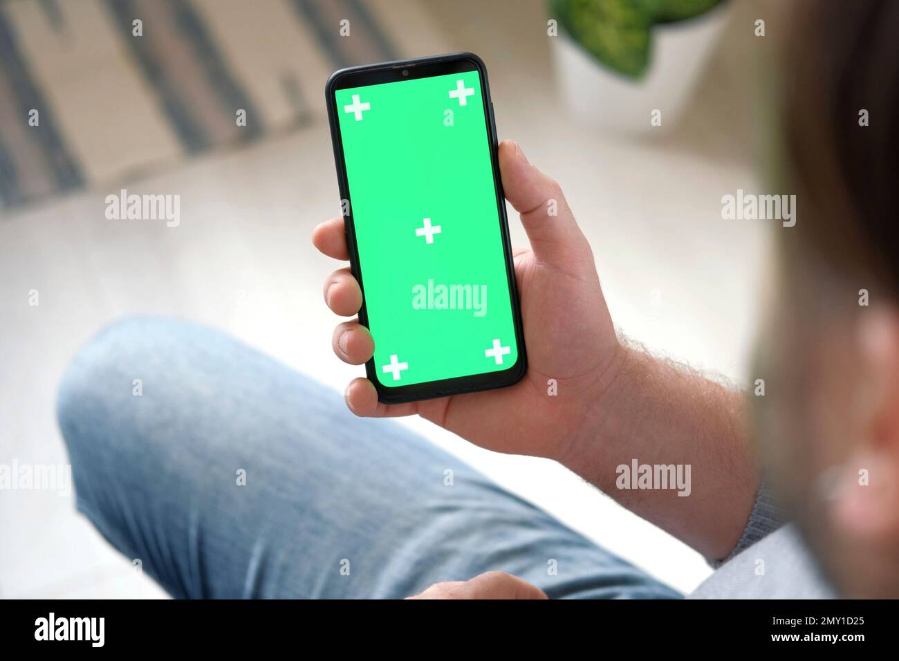 Young man sitting at home holding smartphone green mock-up screen in hand. Male person using ...