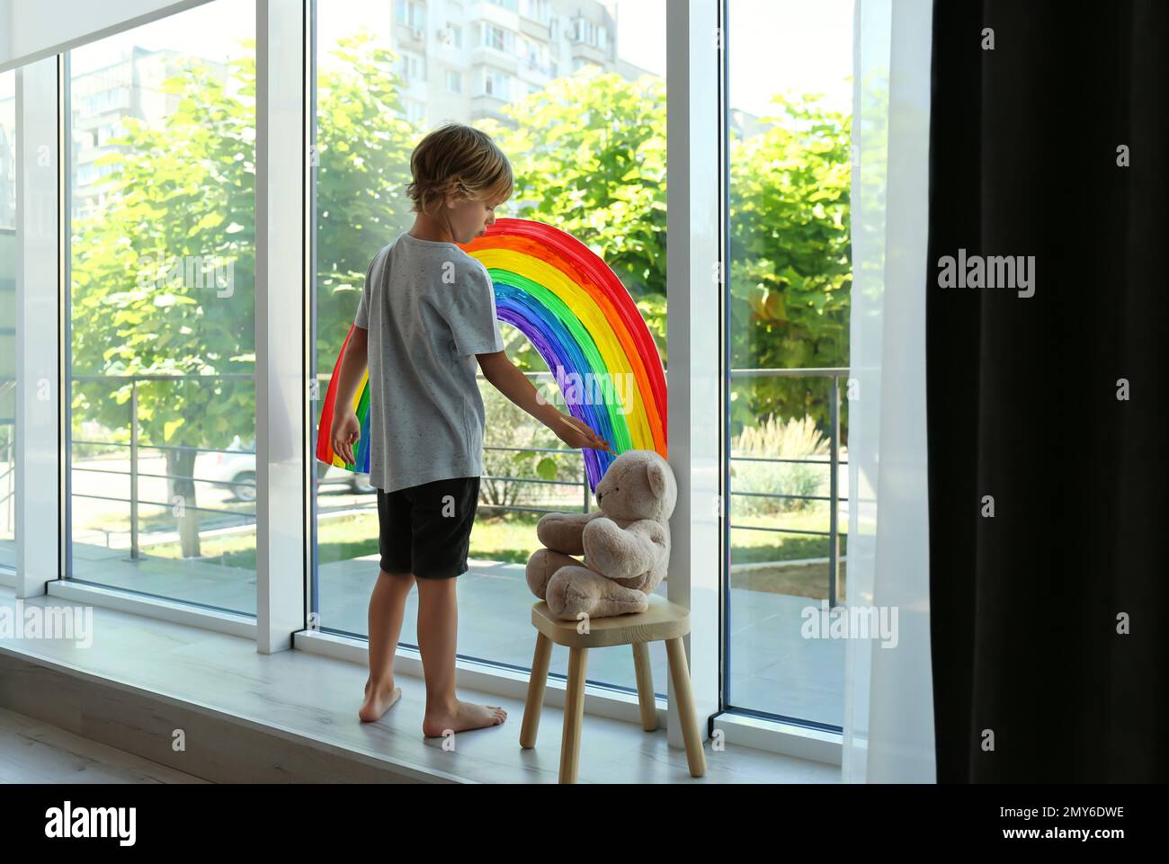 Little boy drawing rainbow on window indoors. Stay at home concept ...