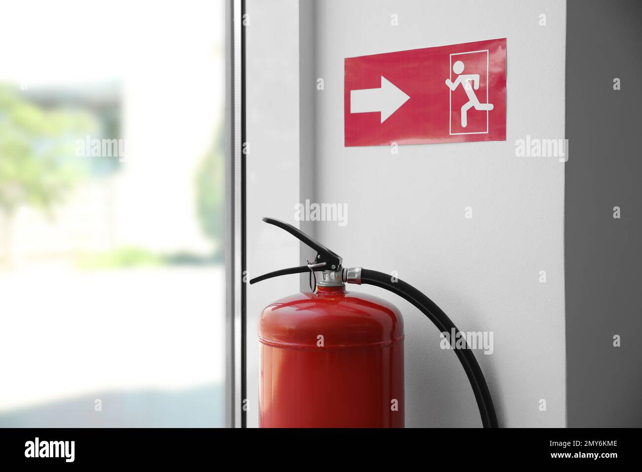 Modern fire extinguisher and emergency exit sign near window indoors ...