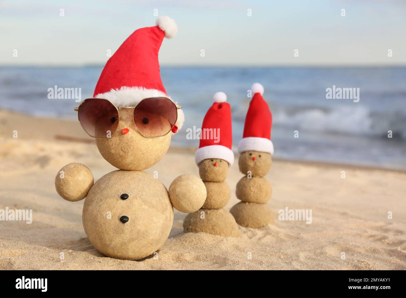 Snowmen made of sand with Santa hats and sunglasses on beach near sea ...