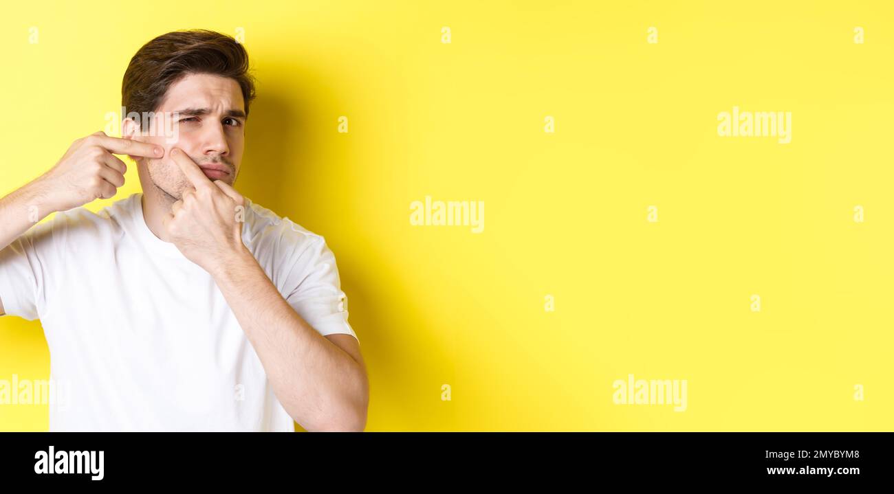 Young man pop a pimple on cheek, standing over yellow background ...