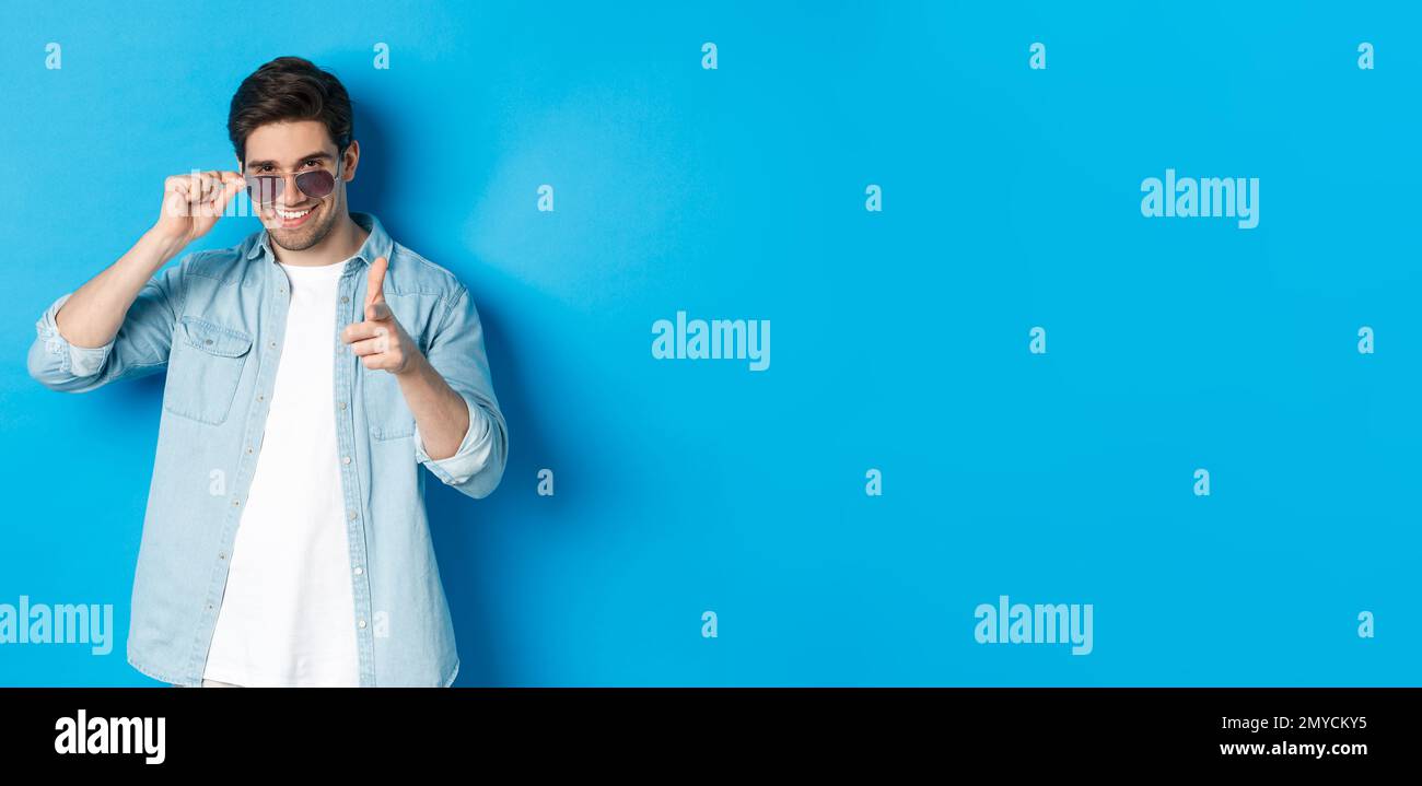Handsome cheeky guy in sunglasses, smiling and pointing finger gun at ...