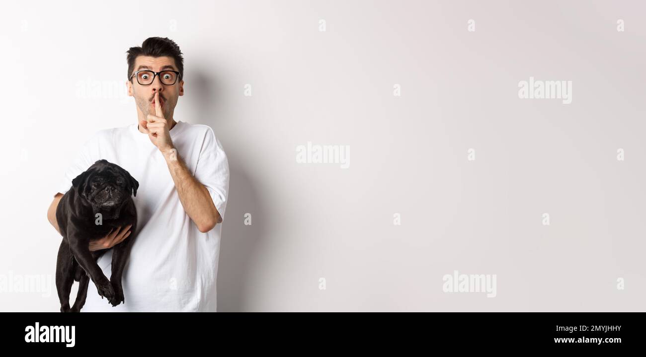 Image of funny young man holding cute black pug dog and shushing at ...