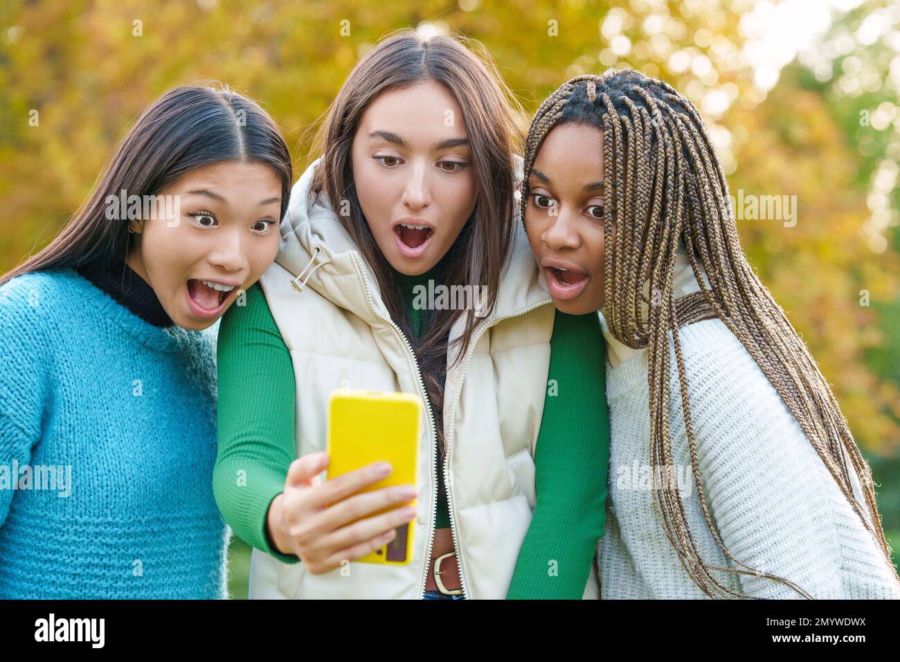 Three surprised friends using a mobile outdoors Stock Photo - Alamy