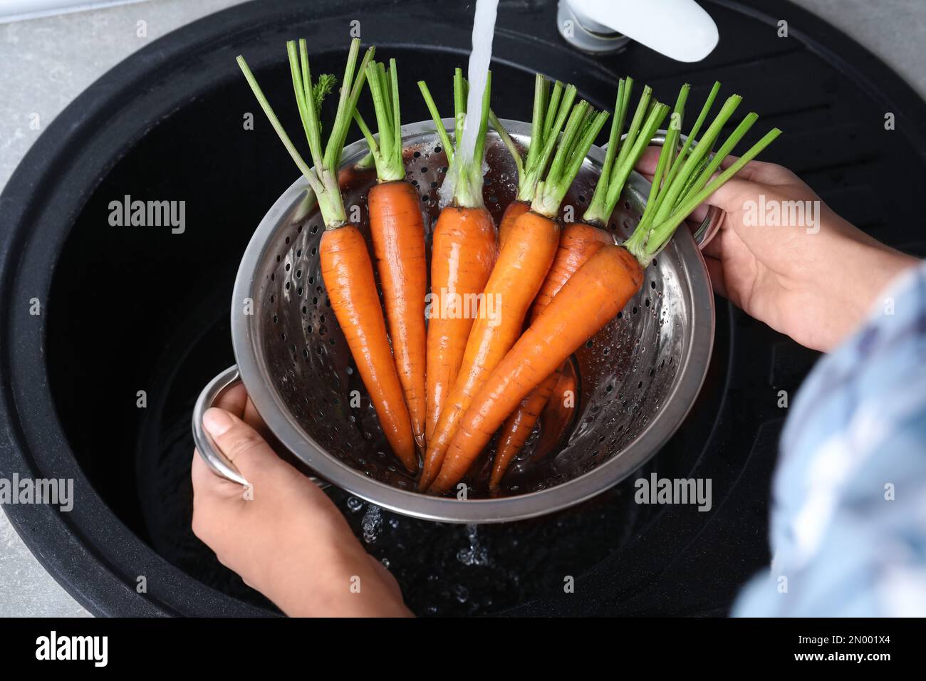 Woman washing ripe carrots in colander with running water over sink ...