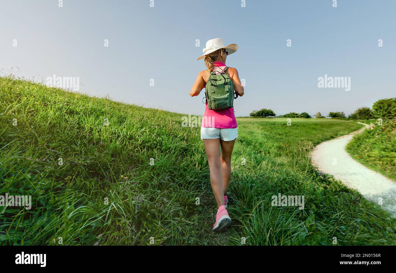a female hiker with her back turned, walks through a green meadow ...