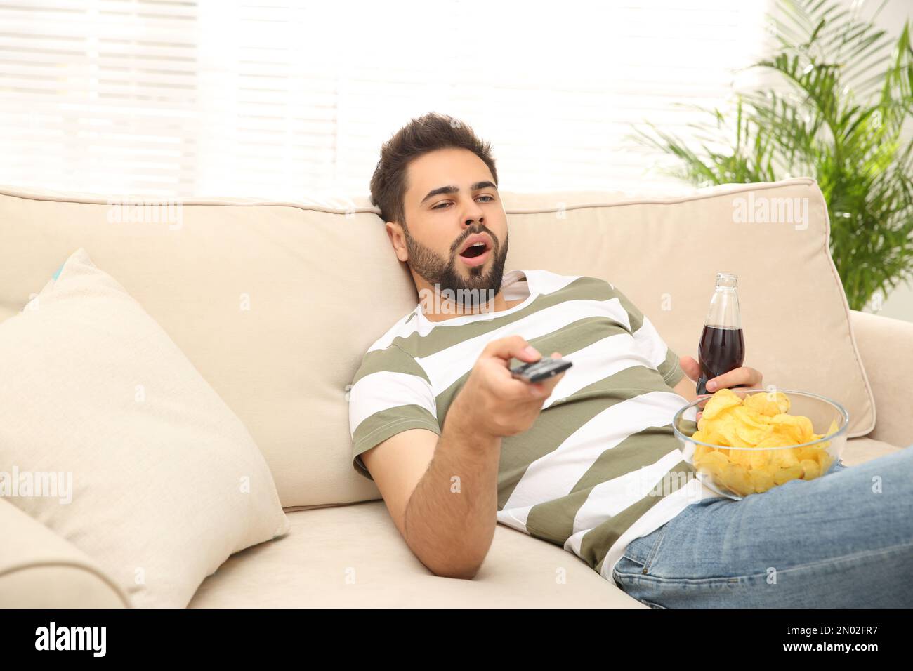 Lazy young man with chips and drink watching TV on sofa at home Stock ...