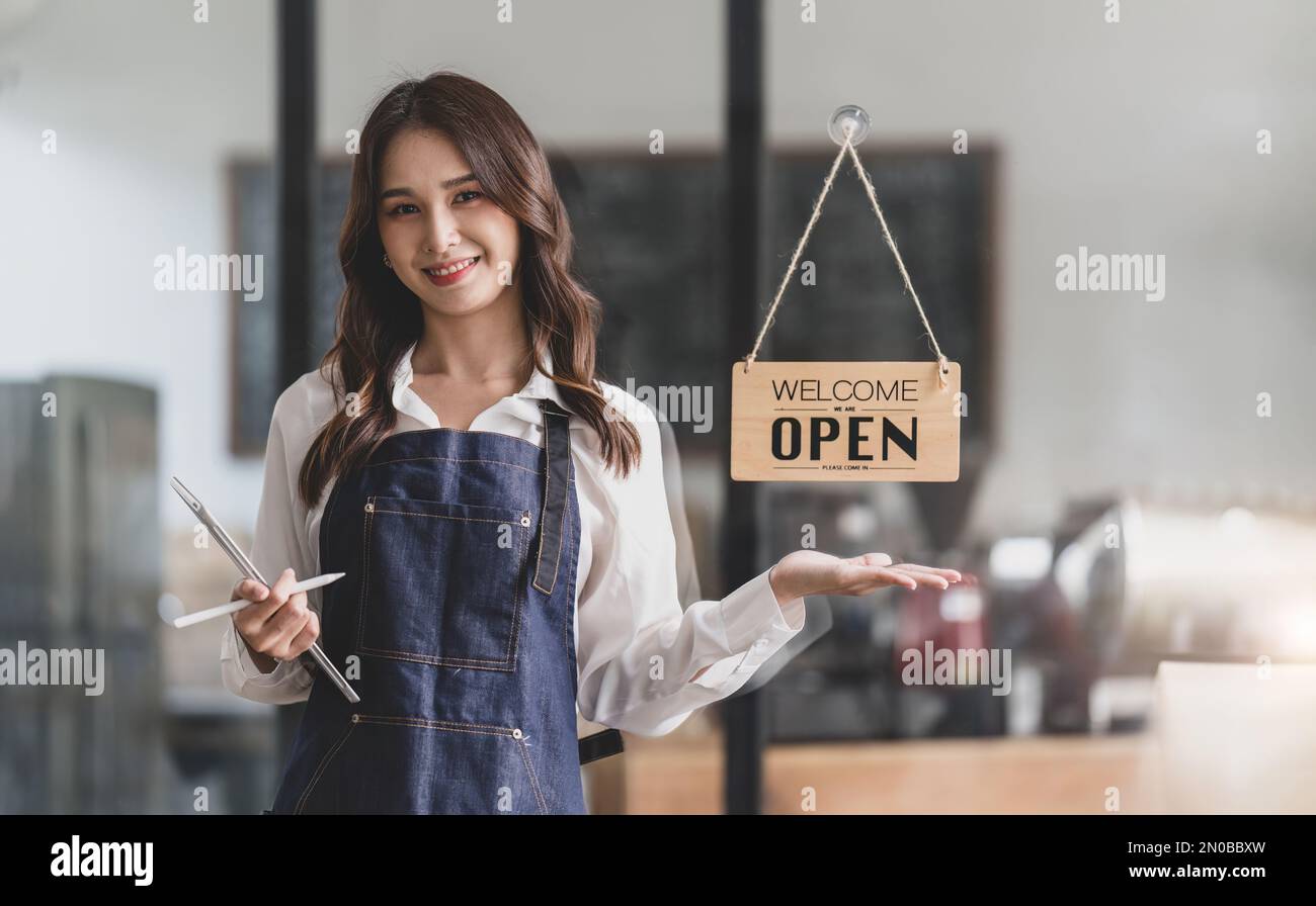 Beautiful asian young barista woman in apron holding tablet and ...