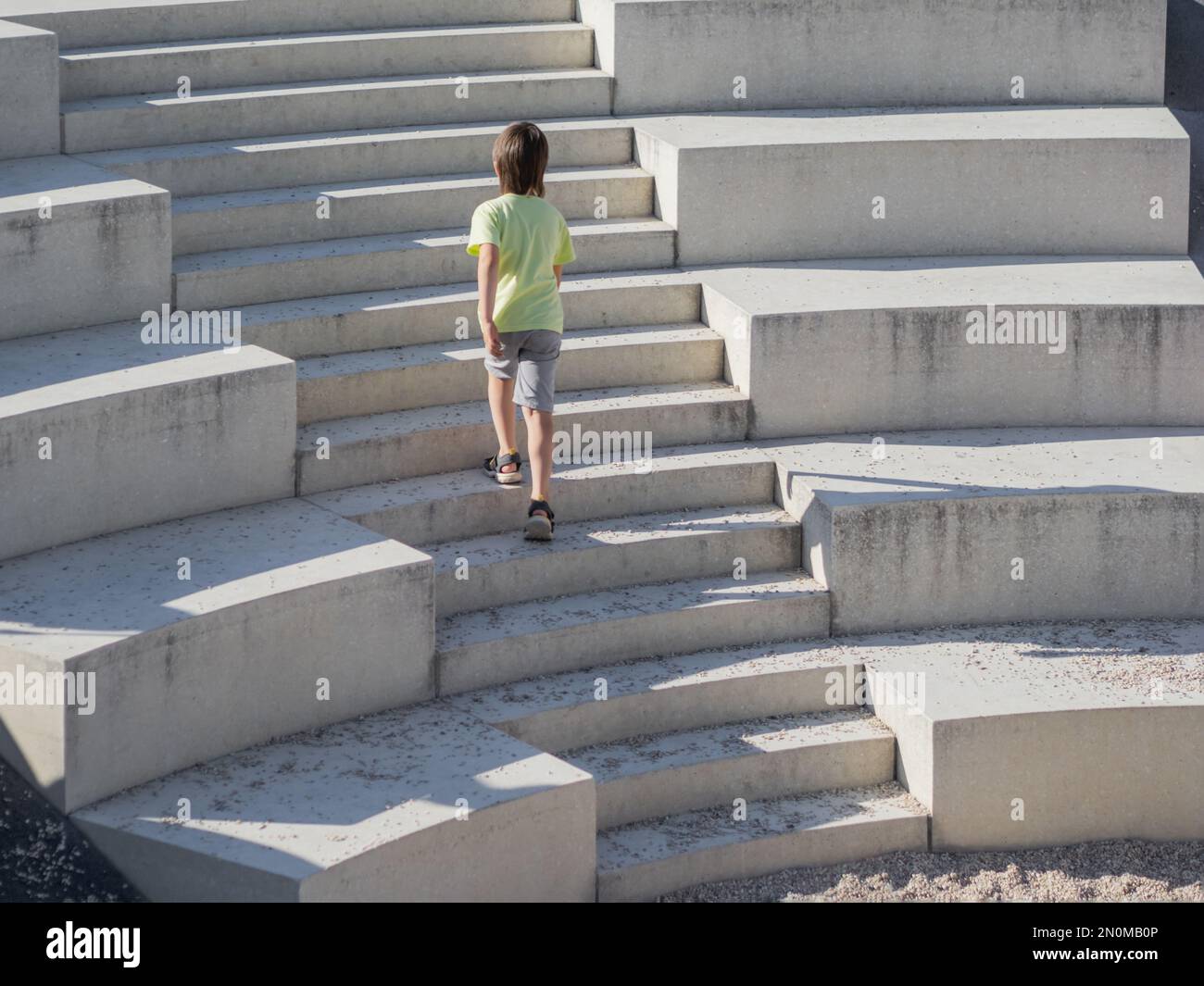 Rear view of boy climbing up the sun-drenched concrete stairs. Kid is ...