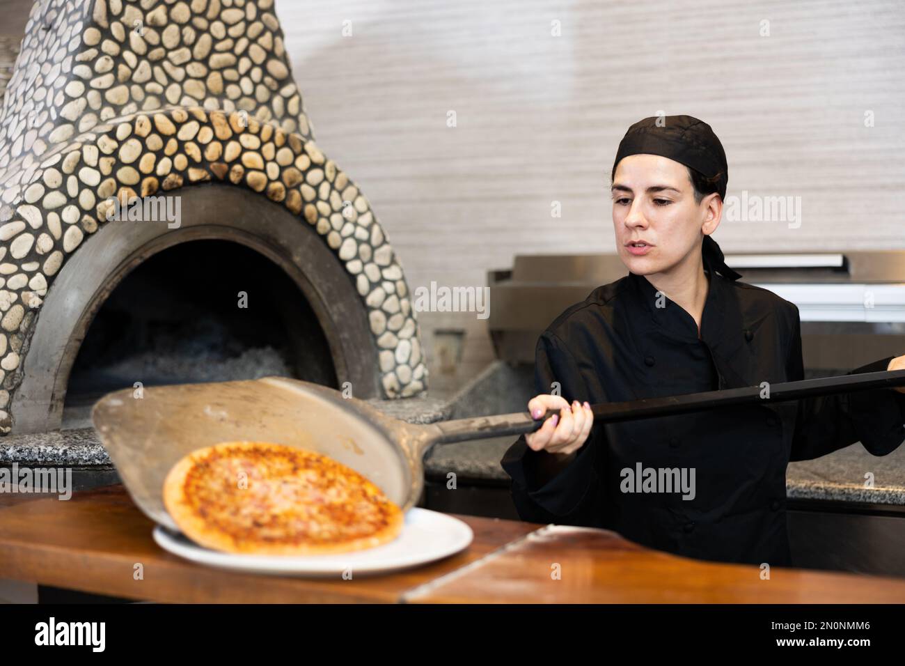 Female chef in black uniform prepares pizza and pulls it out of oven in ...