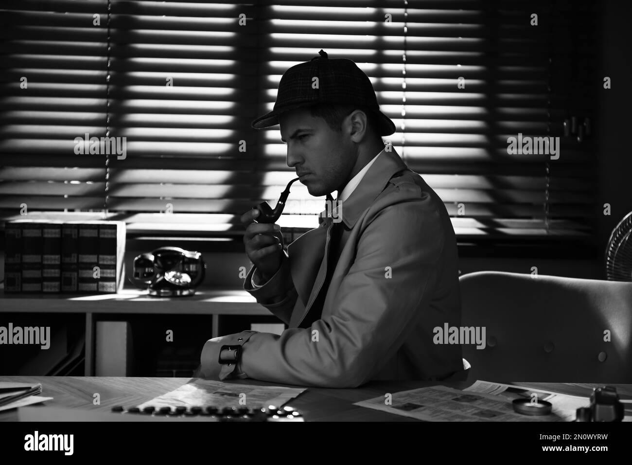 Old fashioned detective with smoking pipe at table in office. Black and ...