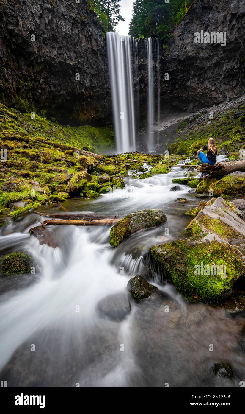 Young woman sitting on riverbank, waterfall cascading over ledge ...