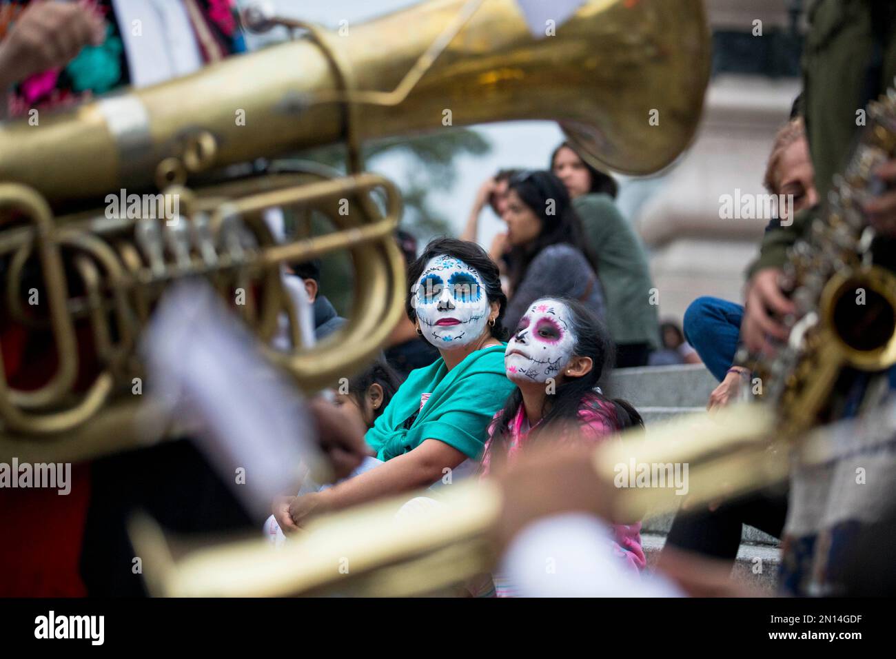 A woman and a girl with their faces painted as "Catrinas," listen to a ...