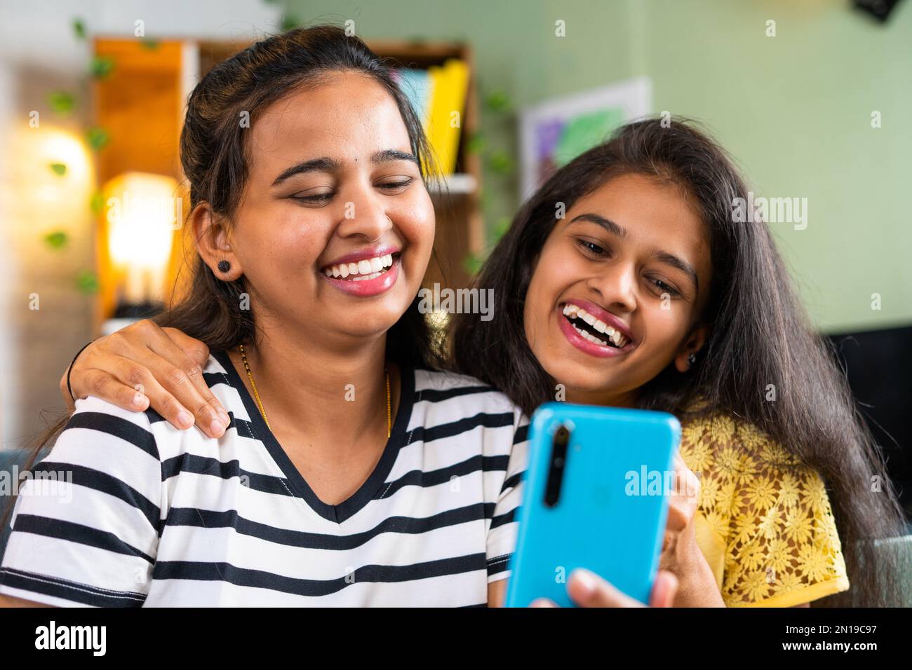 happy sibling girls laughing while using mobile phone on sofa at home ...