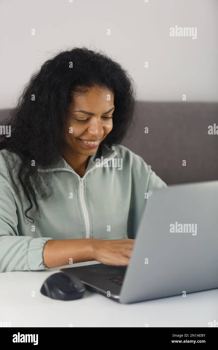 Happy African woman working on modern laptop computer at home. Cheerful BIPOC female typing text ...