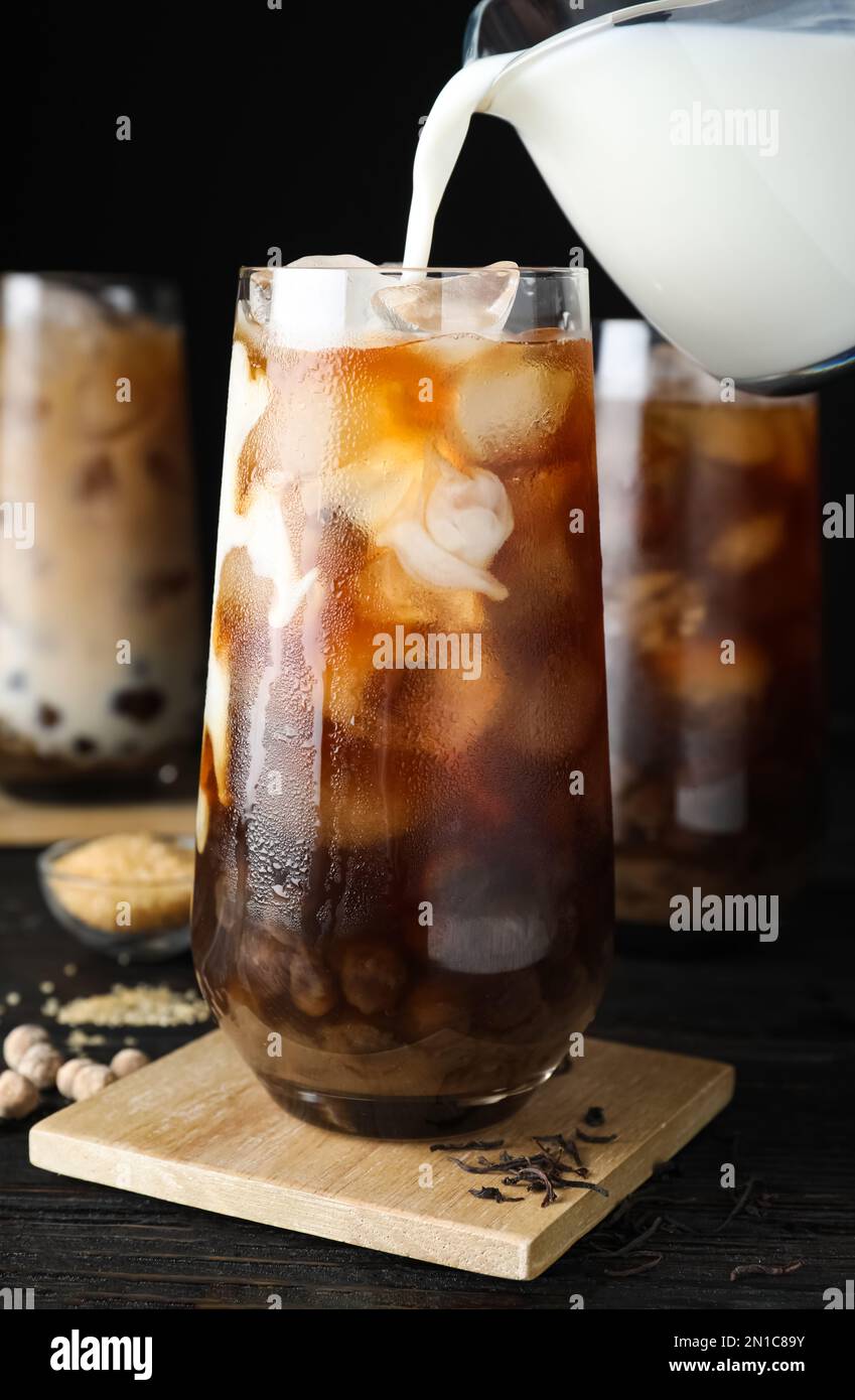 Pouring milk into glass with bubble tea on black wooden table, closeup ...