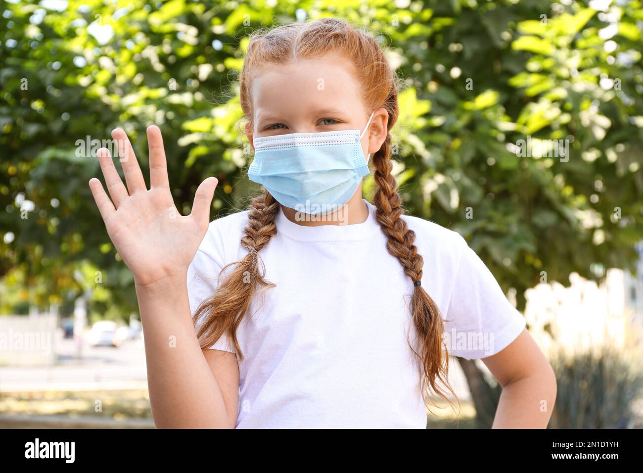 Little girl in protective mask showing hello gesture outdoors. Keeping ...