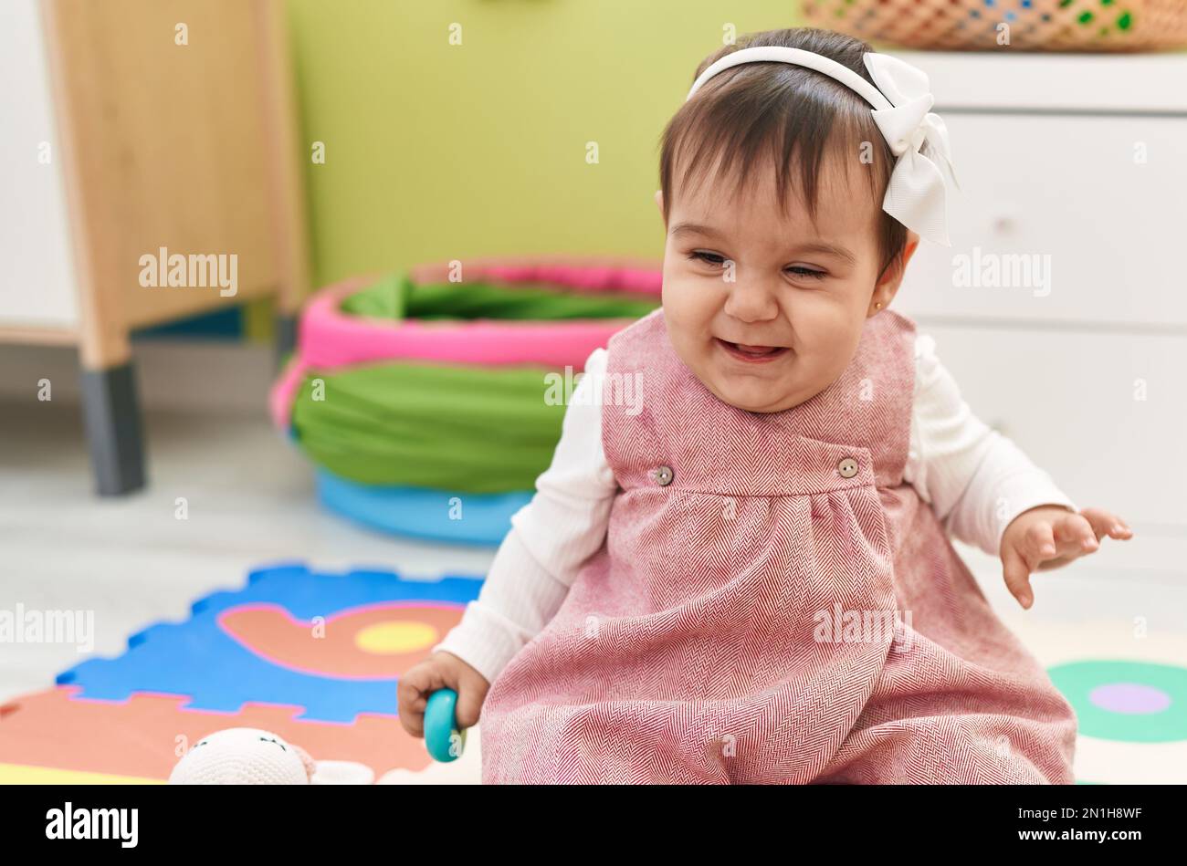 Adorable hispanic baby sitting on floor holding toy at kindergarten ...