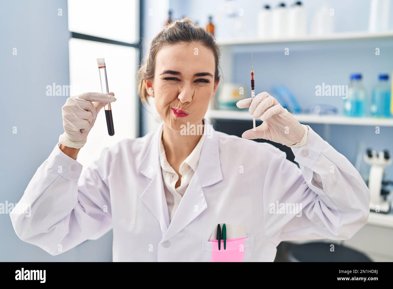 Young woman working at scientist laboratory holding blood sample puffing cheeks with funny face ...