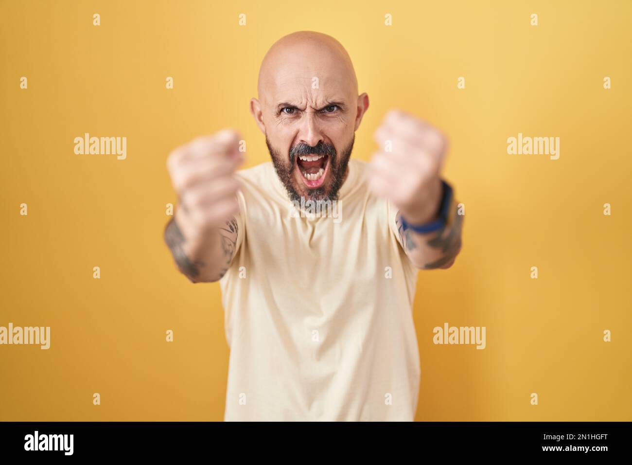 Hispanic man with tattoos standing over yellow background angry and mad ...