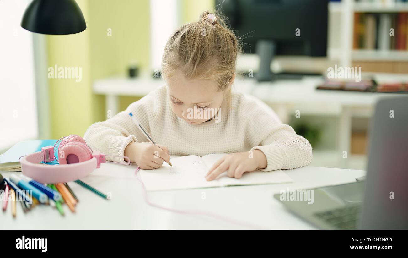 Adorable blonde girl student writing on notebook sitting on table at ...