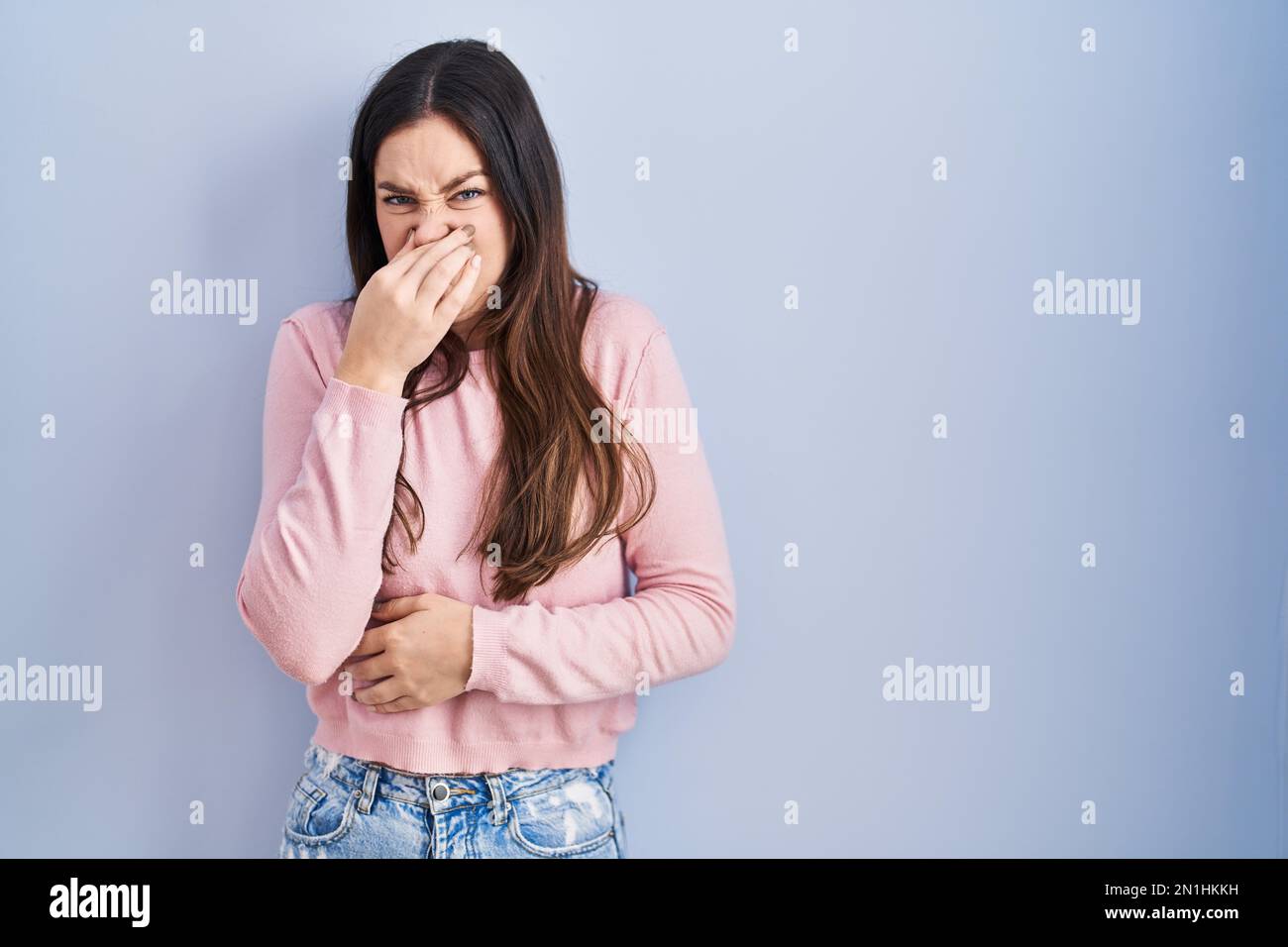 Young brunette woman standing over blue background smelling something ...