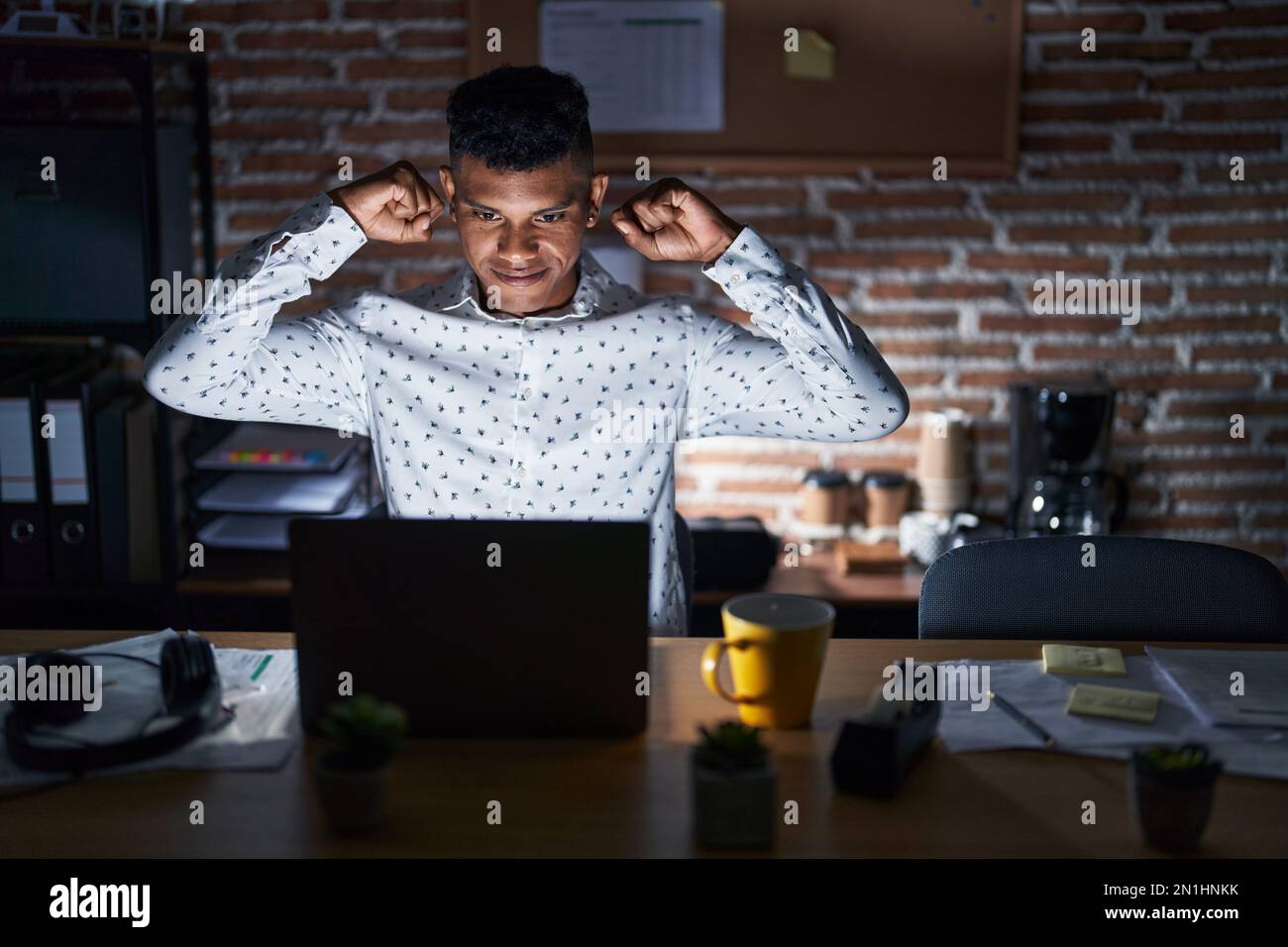 Young hispanic man working at the office at night showing arms muscles ...