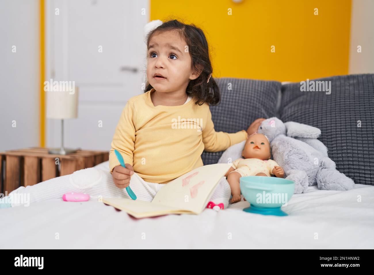Adorable hispanic toddler sitting on bed reading book at bedroom Stock ...