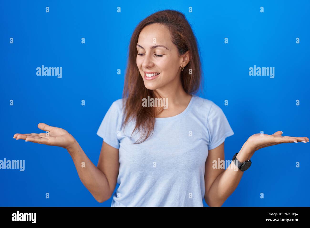 Brunette woman standing over blue background smiling showing both hands ...