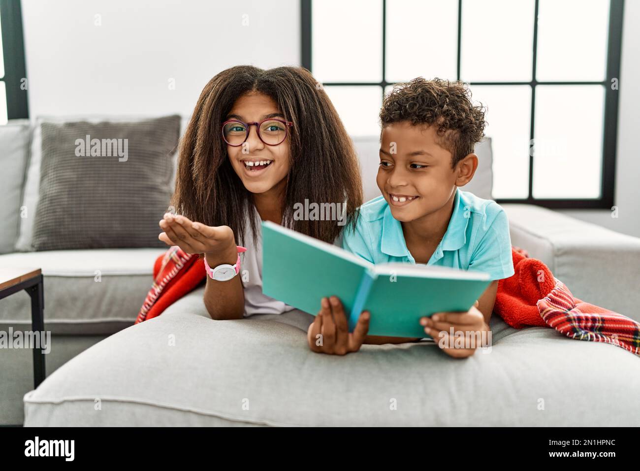 Two siblings lying on the sofa reading a book pointing aside with hands ...