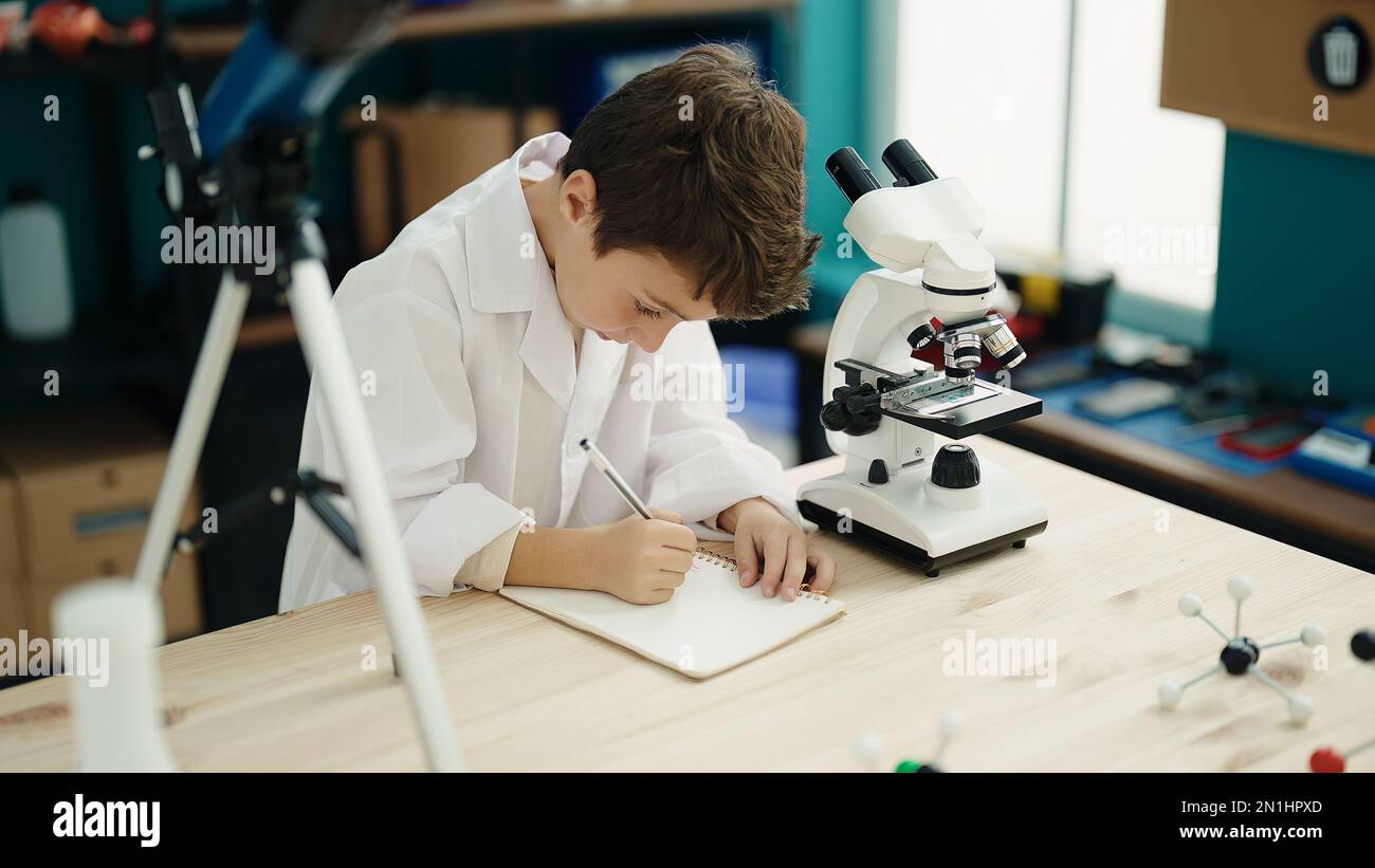 Adorable hispanic boy student using microscope writing on notebook at ...