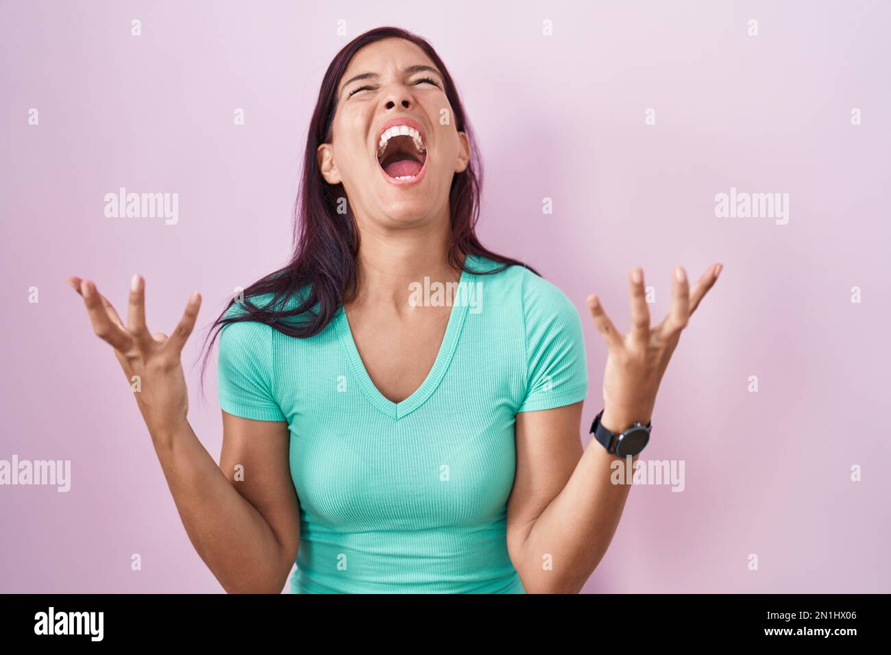 Young hispanic woman standing over pink background crazy and mad ...