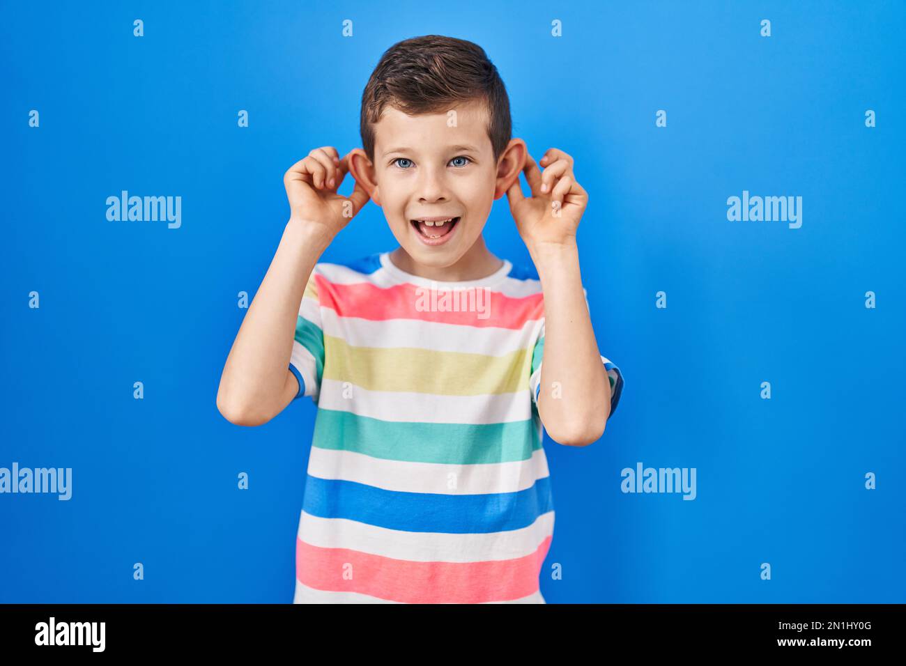 Young caucasian kid standing over blue background smiling pulling ears ...