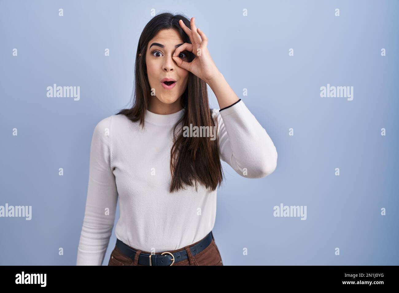 Young brunette woman standing over blue background doing ok gesture ...