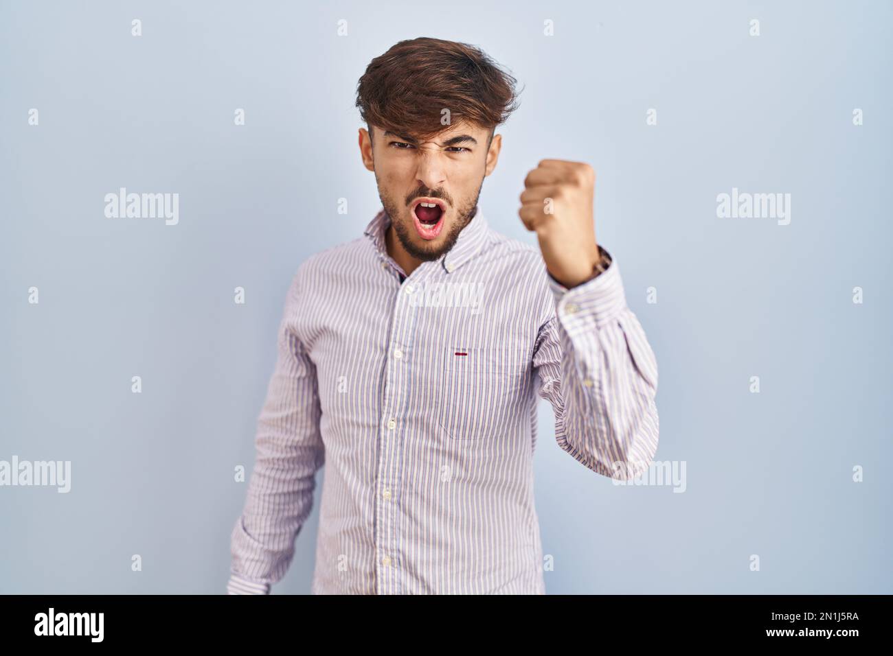 Arab man with beard standing over blue background angry and mad raising ...