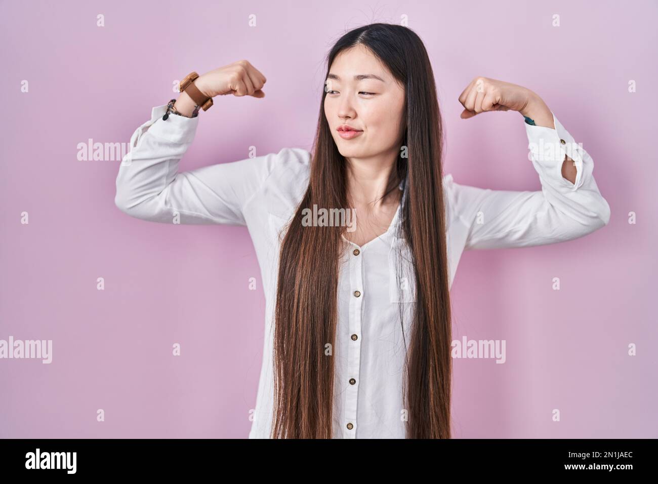 Chinese young woman standing over pink background showing arms muscles ...