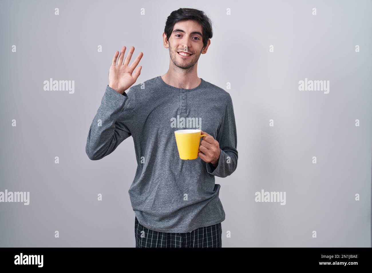 Young hispanic man wearing pajama drinking a cup of coffee waiving ...