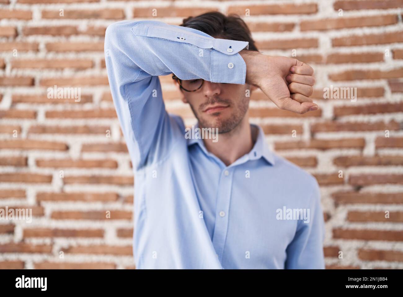 Young hispanic man standing over brick wall background covering eyes ...