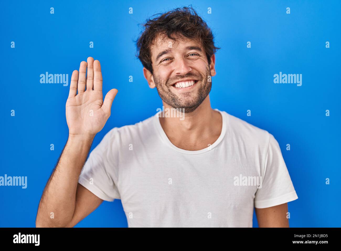 Hispanic young man standing over blue background waiving saying hello ...