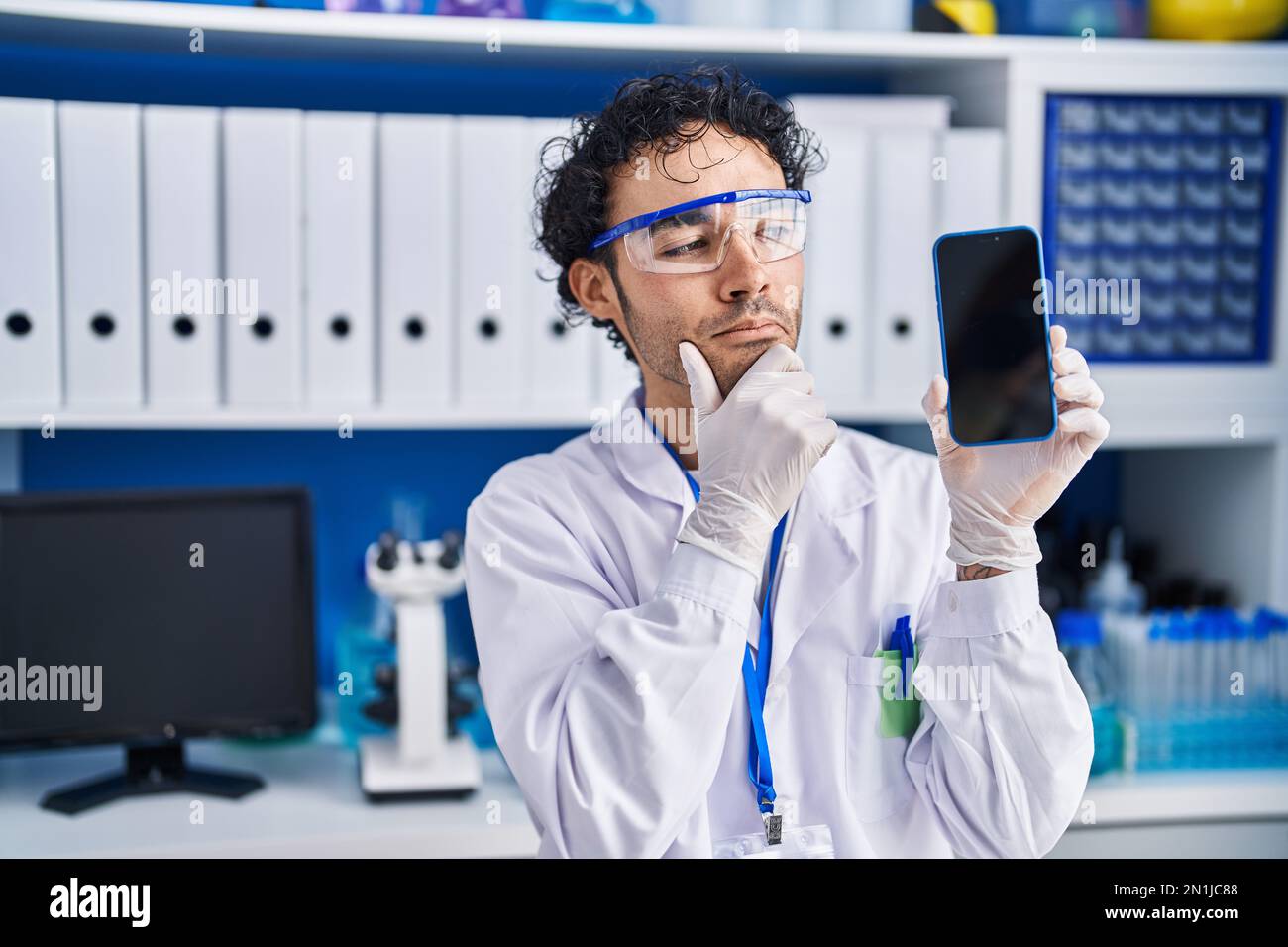 Hispanic man working at scientist laboratory showing smartphone screen serious face thinking ...