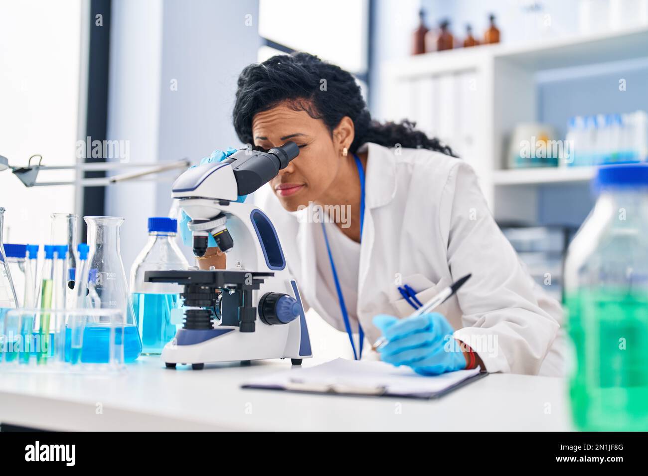 African american woman wearing scientist uniform using microscope write on document at ...