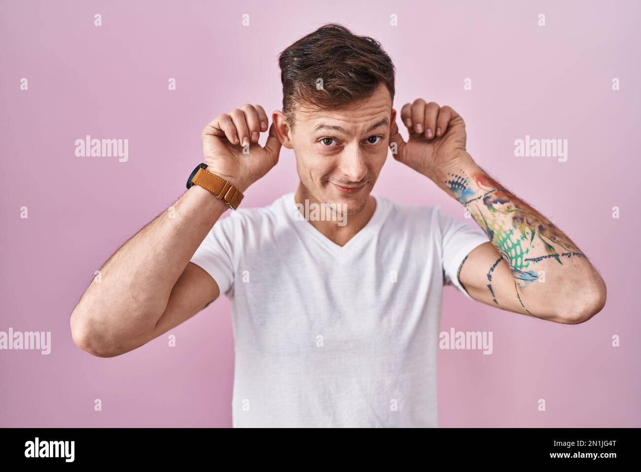 Caucasian man standing over pink background smiling pulling ears with ...
