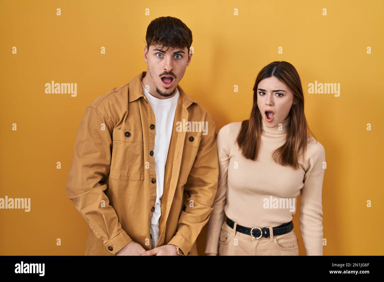 Young hispanic couple standing over yellow background in shock face ...