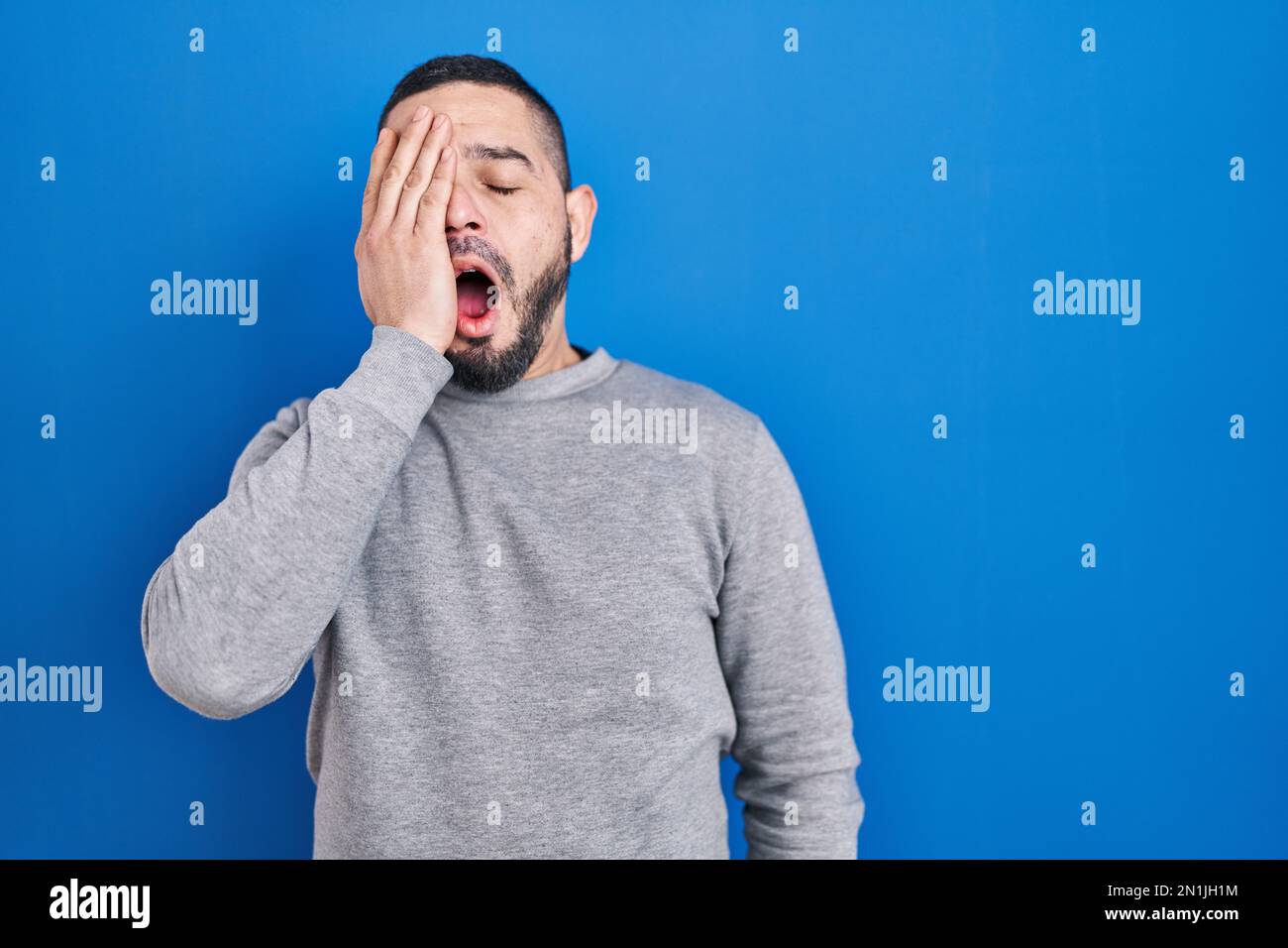 Hispanic man standing over blue background yawning tired covering half ...