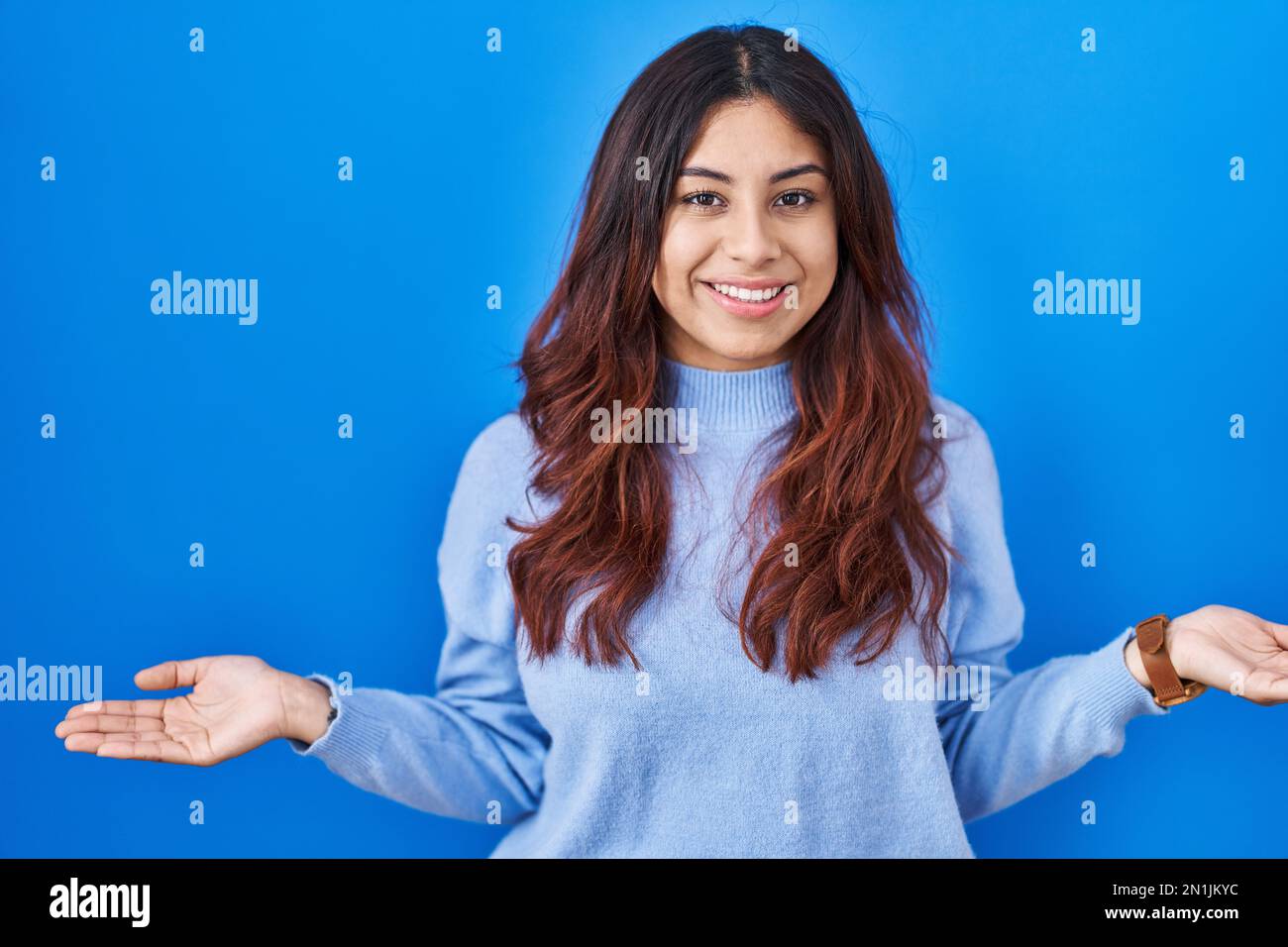 Hispanic young woman standing over blue background clueless and ...