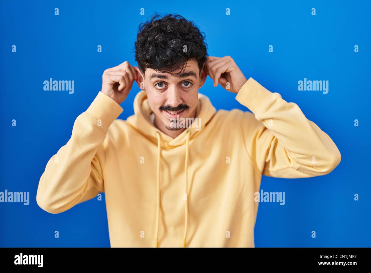 Hispanic man standing over blue background smiling pulling ears with ...