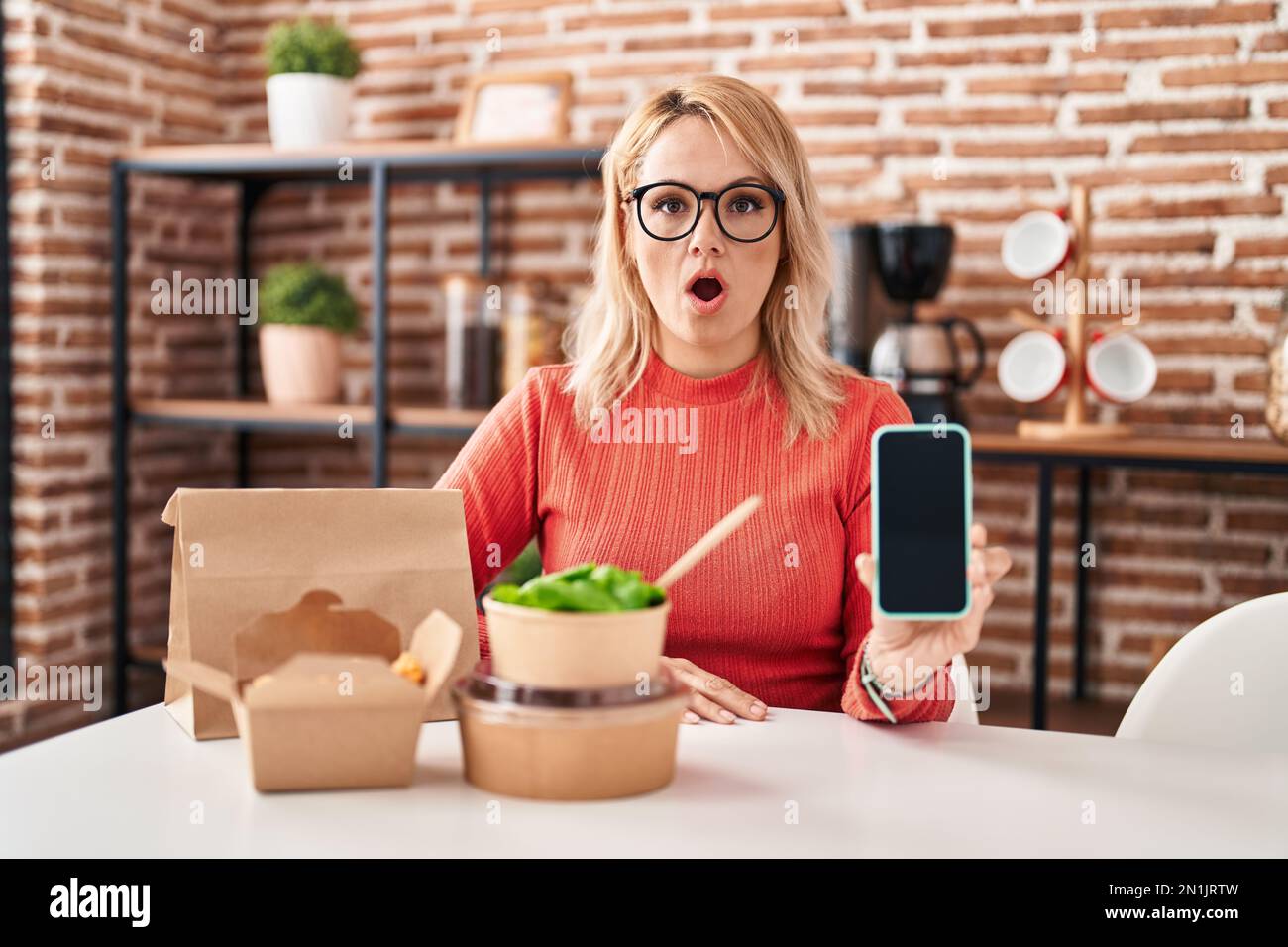 Blonde woman eating take away food showing smartphone screen scared and ...