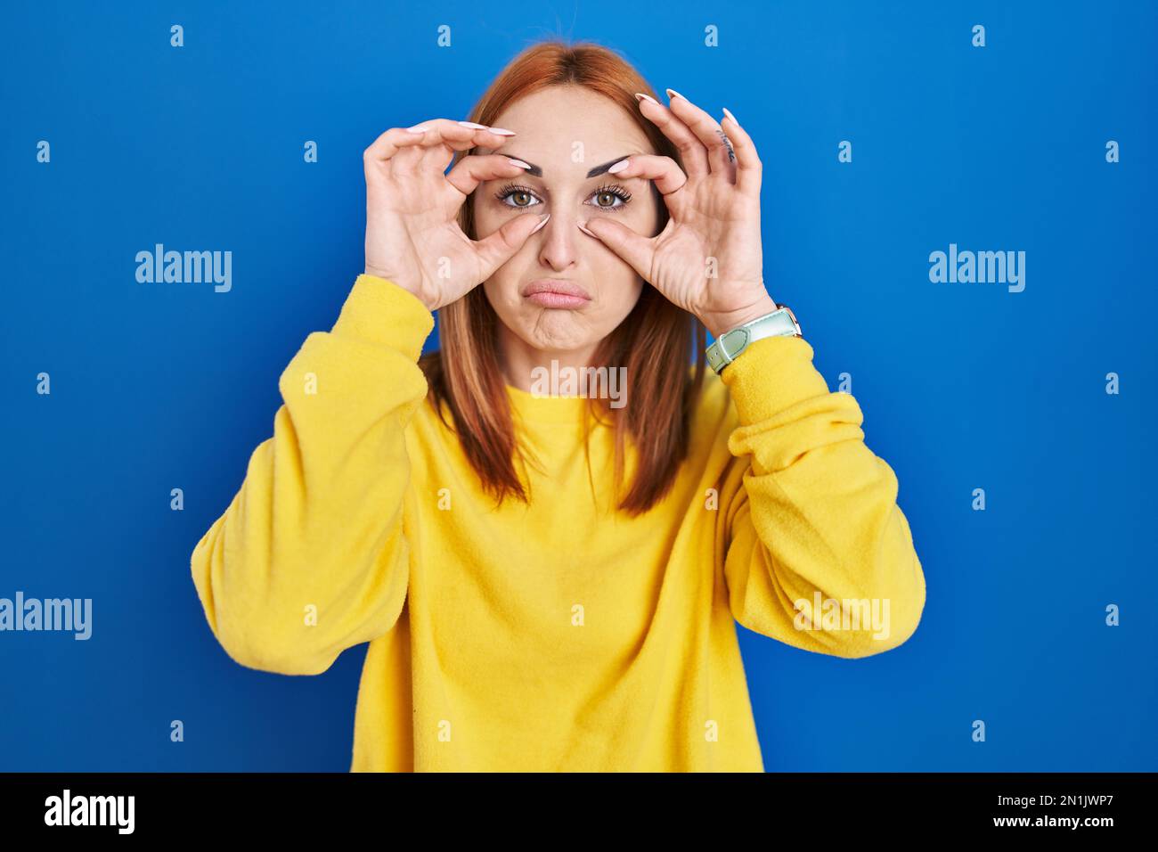 Young woman standing over blue background trying to open eyes with ...