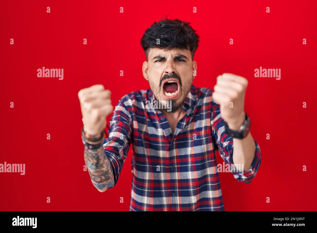 Young hispanic man with beard standing over red background angry and ...