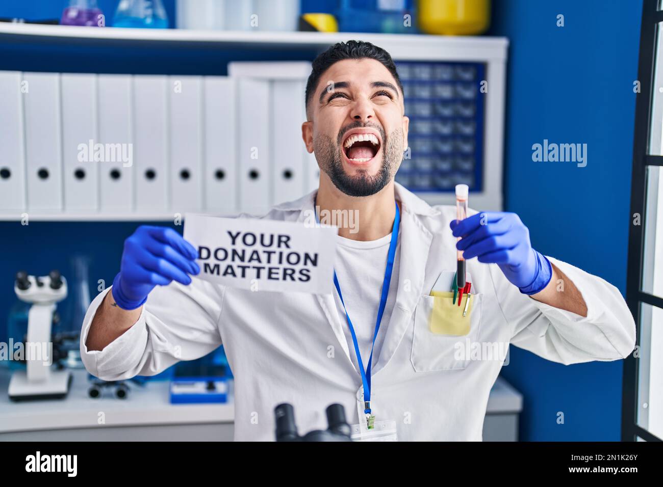 Young handsome man working at scientist laboratory holding blood sample angry and mad screaming ...